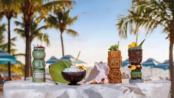 Uma mesa com quatro bebidas estilo tiki, um coco e uma concha, com o cenário de uma praia e palmeiras ao fundo.