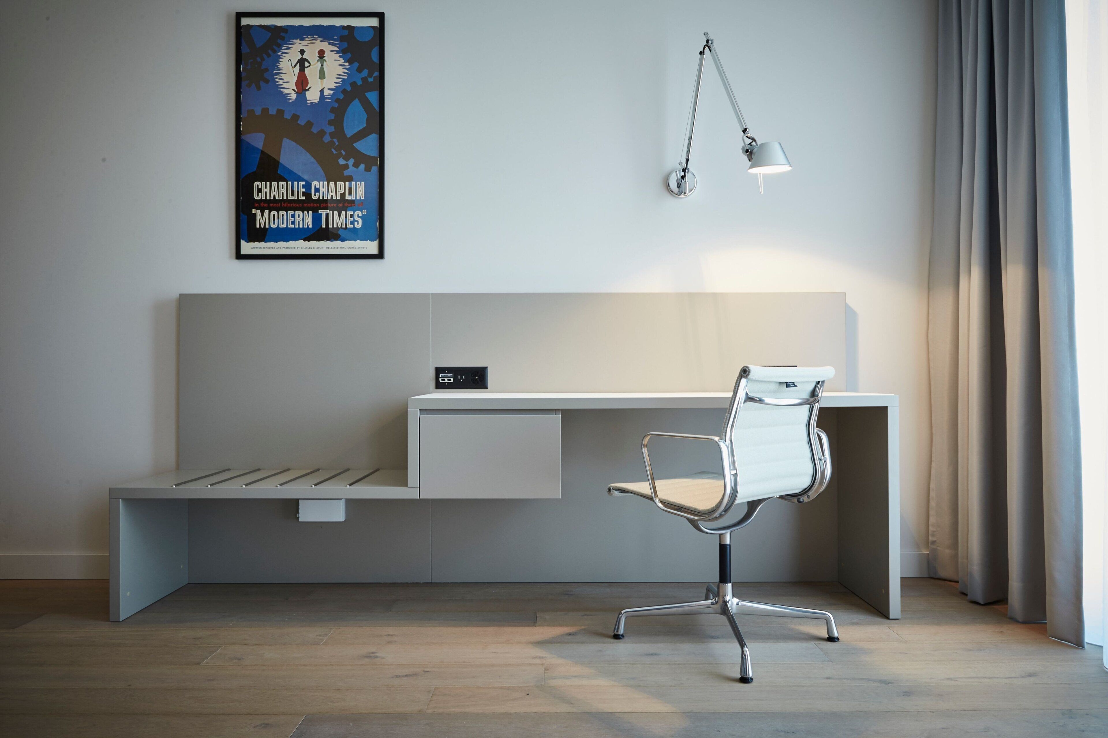 Um espaço de escritório moderno, com uma mesa branca, uma cadeira de metal, um cartaz na parede e uma luminária.