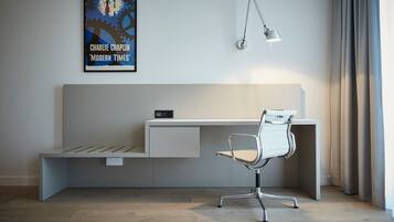 Um espaço de escritório moderno, com uma mesa branca, uma cadeira de metal, um cartaz na parede e uma luminária.