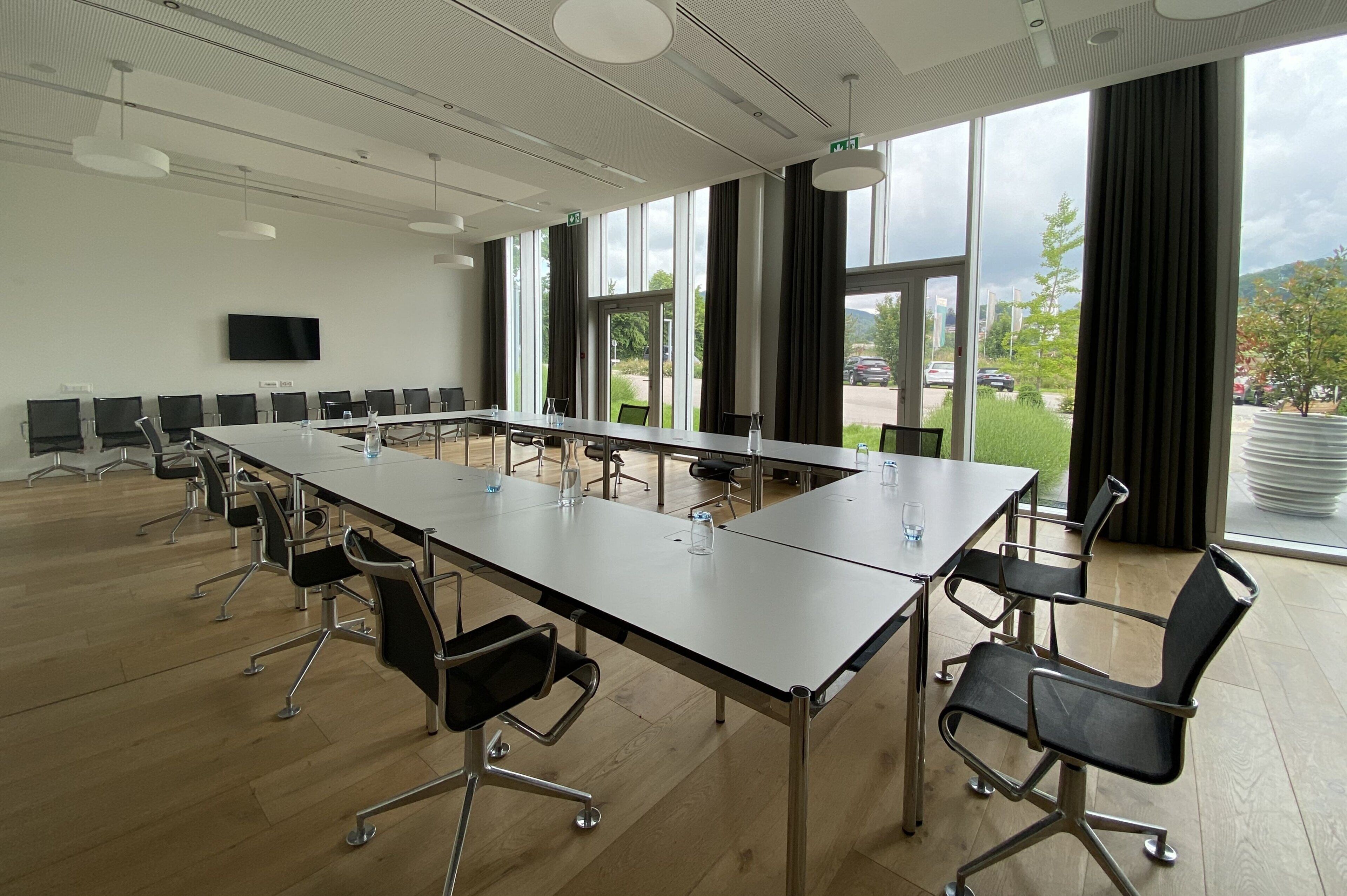 Uma sala de conferências com uma mesa comprida, cadeiras, uma TV de tela plana e janelas grandes com cortinas.