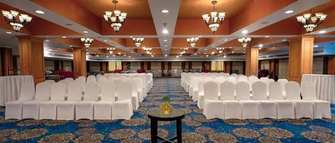 Uma sala de banquetes com fileiras de cadeiras brancas, um tapete azul com padrão floral e lustres no teto.