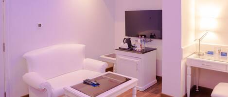 Quarto de hotel com poltrona branca, mesa pequena com controle remoto, minibar, escrivaninha com luminária e espelho.