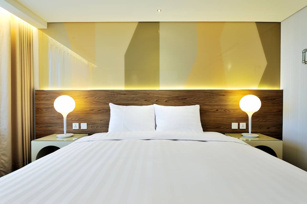 Cama bem arrumada com lençóis brancos, duas luminárias de cabeceira e cabeceira de madeira.
