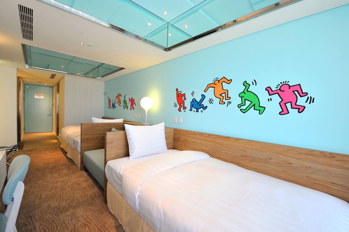 Quarto de hotel com uma cama grande, uma cama menor, uma escrivaninha e uma cadeira. Há uma parede com arte colorida e um teto com claraboia.