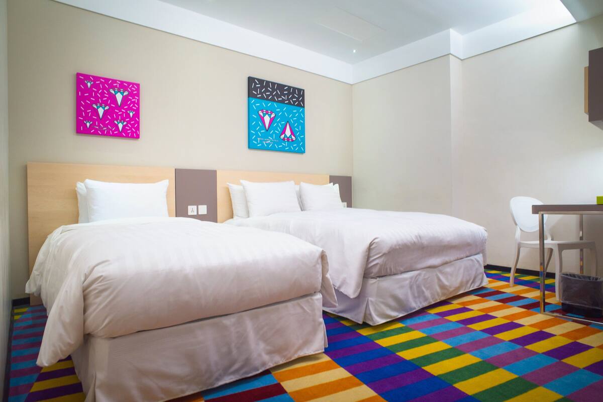 Quarto de hotel com duas camas, uma escrivaninha e carpete listrado colorido.