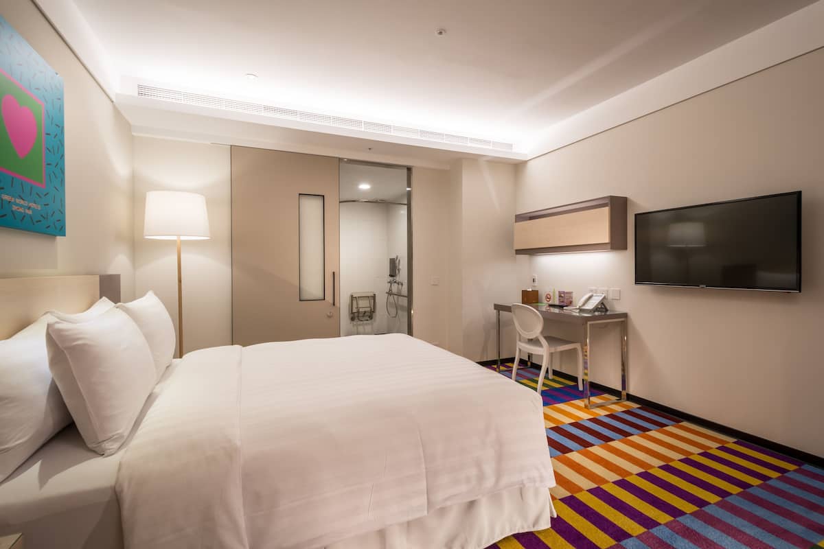 Quarto de hotel com cama, escrivaninha, cadeira, televisão e um tapete listrado e colorido.