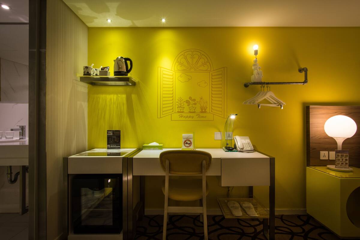 Quarto de hotel com escrivaninha, cadeira e prateleira na parede com chaleira e caixa de lenços.