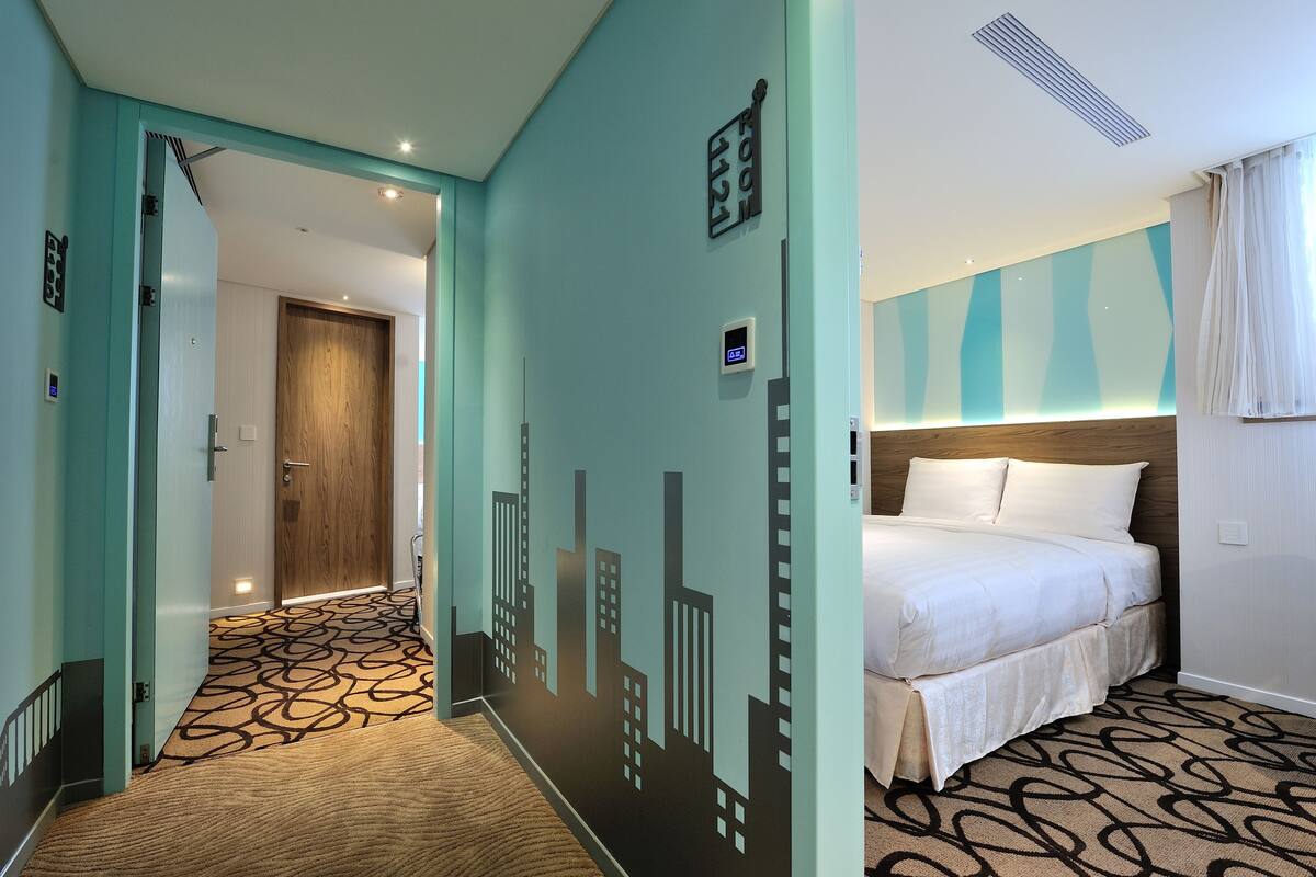 Quarto de hotel com cama, um mural de paisagem urbana, uma porta e um corredor com carpete.