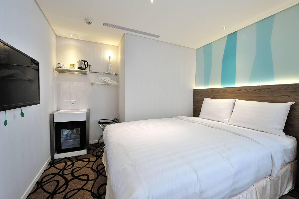 Quarto de hotel com uma cama grande, um espaço de estar pequeno com um aquecedor, uma televisão e uma prateleira na parede com uma chaleira.