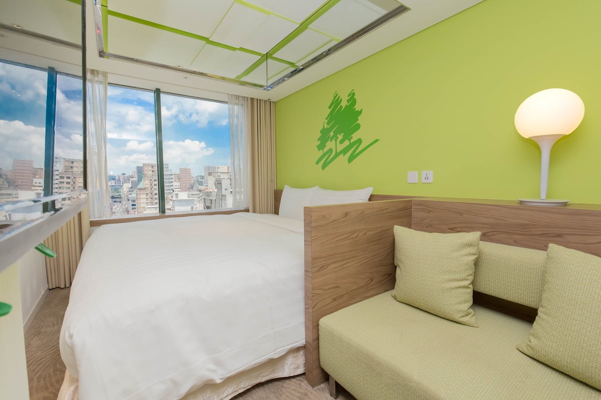 Quarto de hotel com uma cama grande, um sofá, vista para a cidade e parede verde com um detalhe decorativo.