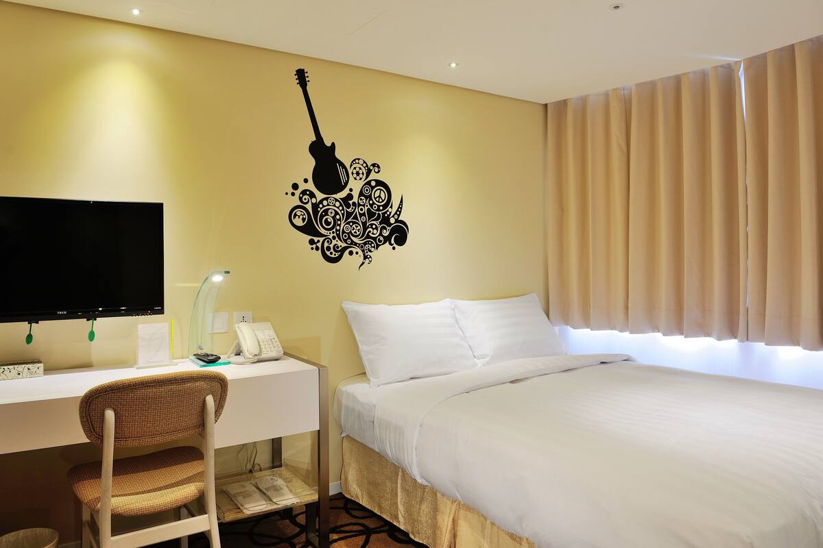 Quarto de hotel com cama, escrivaninha com monitor de computador, cadeira e um objeto decorativo de guitarra na parede.