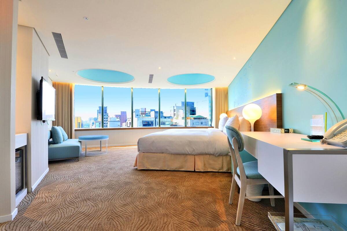 Quarto de hotel com cama, escrivaninha, cadeira, televisão, janela com vista para a cidade e piso coberto de carpete.