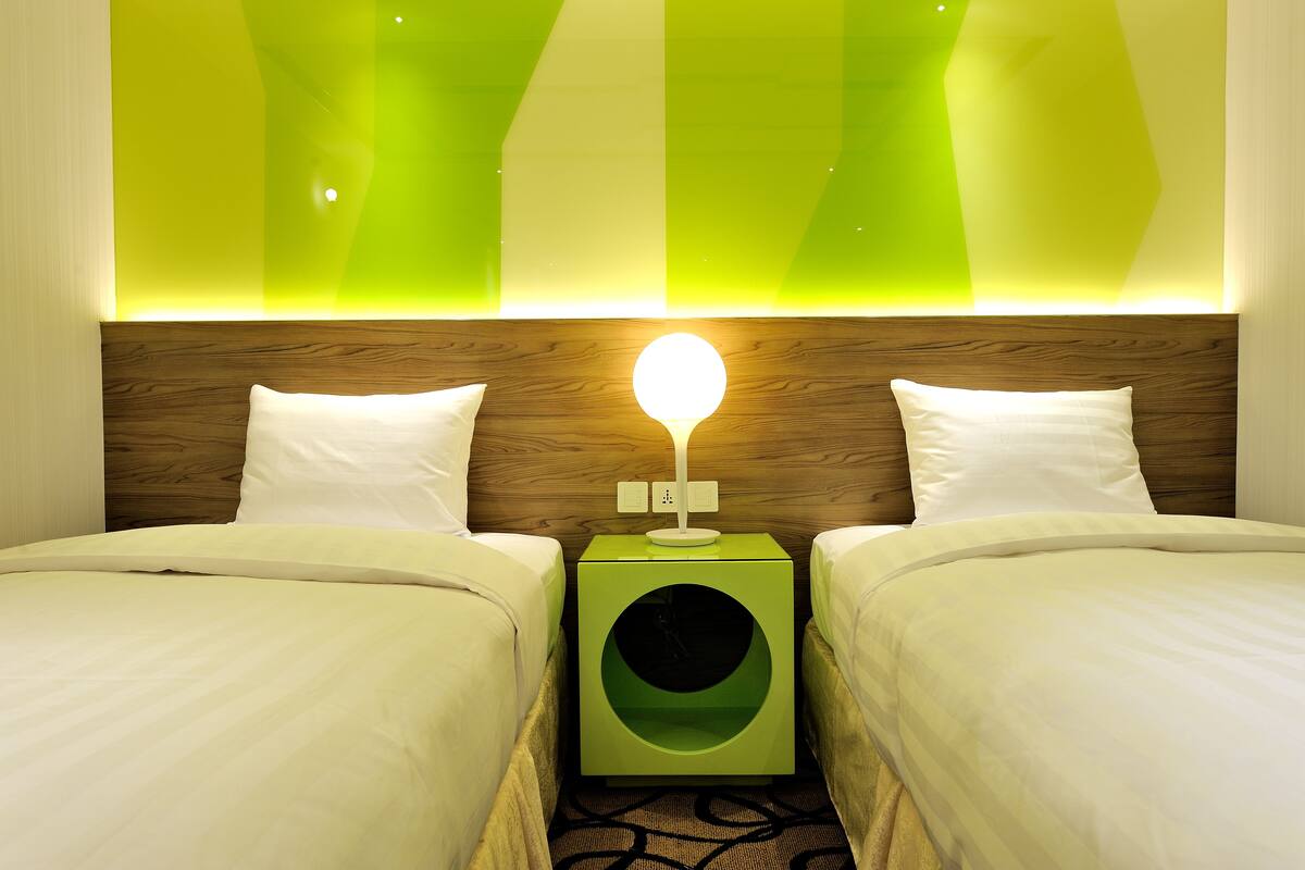 Quarto de hotel com duas camas, uma mesa de cabeceira verde com luminária e um design de teto moderno.