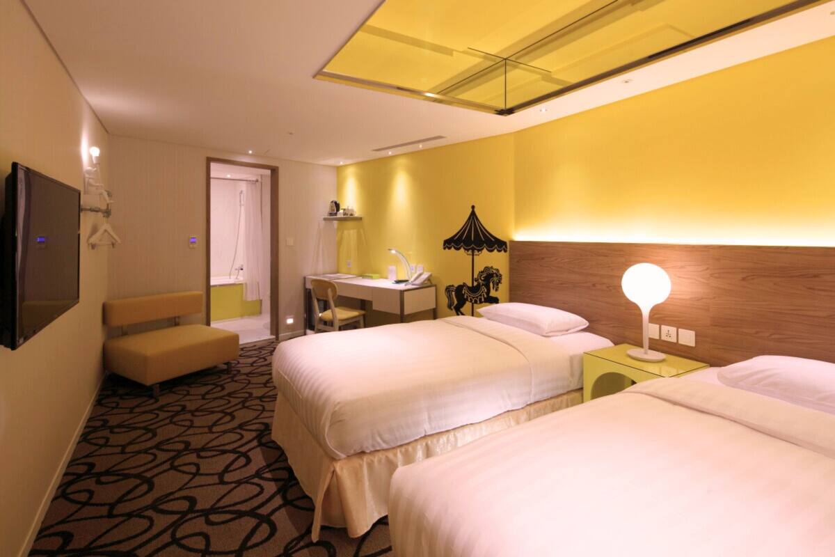 Quarto de hotel com duas camas, uma escrivaninha, uma cadeira, um espaço de estar pequeno e um espelho.