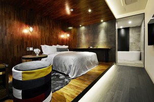 Quarto moderno com teto de madeira, uma cama grande, mesa de cabeceira, um banquinho redondo pequeno e uma banheira embutida.