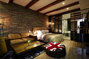 Um quarto moderno com parede de tijolos, vigas de madeira no teto, uma cama com cabeceira de madeira, uma mesinha de cabeceira, um sofá, uma mesa de centro e uma almofada com a bandeira Union Jack.
