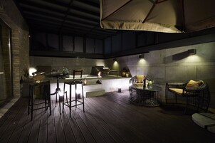 Uma área de varanda externa com piso de madeira, cadeiras de metal, uma mesa redonda e um guarda-sol grande oferecendo sombra.