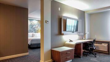Quarto de hotel com cama, escrivaninha com cadeira, televisão presa na parede e termostato na parede.