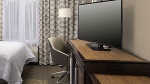 Quarto de hotel com uma cama, uma escrivaninha com televisão, uma cadeira e um abajur.