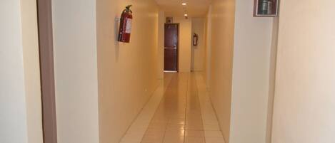 Corredor de hotel com extintor, uma porta no final e um quadro na parede.