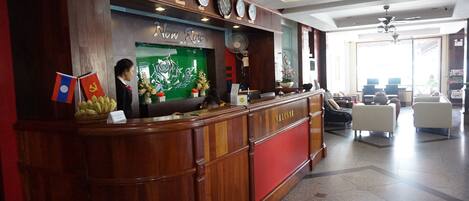 Bancada de recepção com relógios, um letreiro 'New Rose' e um aviso de 'Bem-vindo'. Há bandeiras, uma cesta de frutas e uma parede verde com obras de arte.