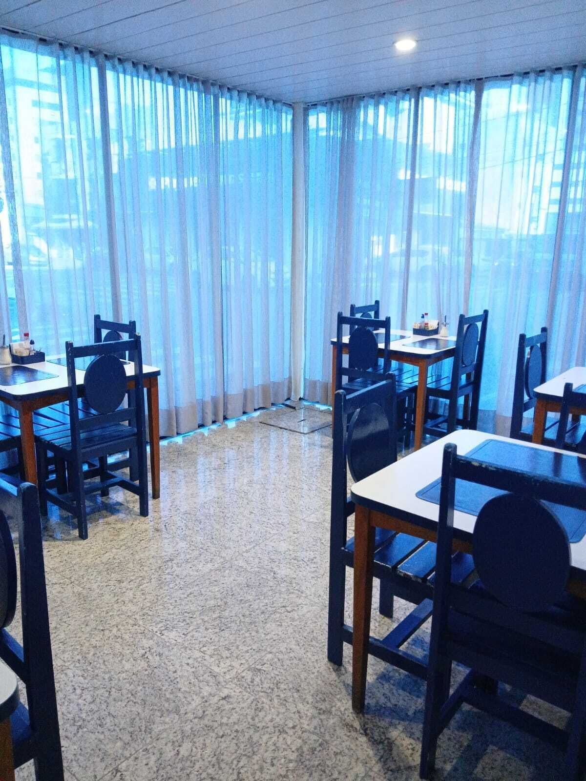 Uma sala de jantar com mesas e cadeiras, janelas amplas com cortinas leves e piso em azulejos.