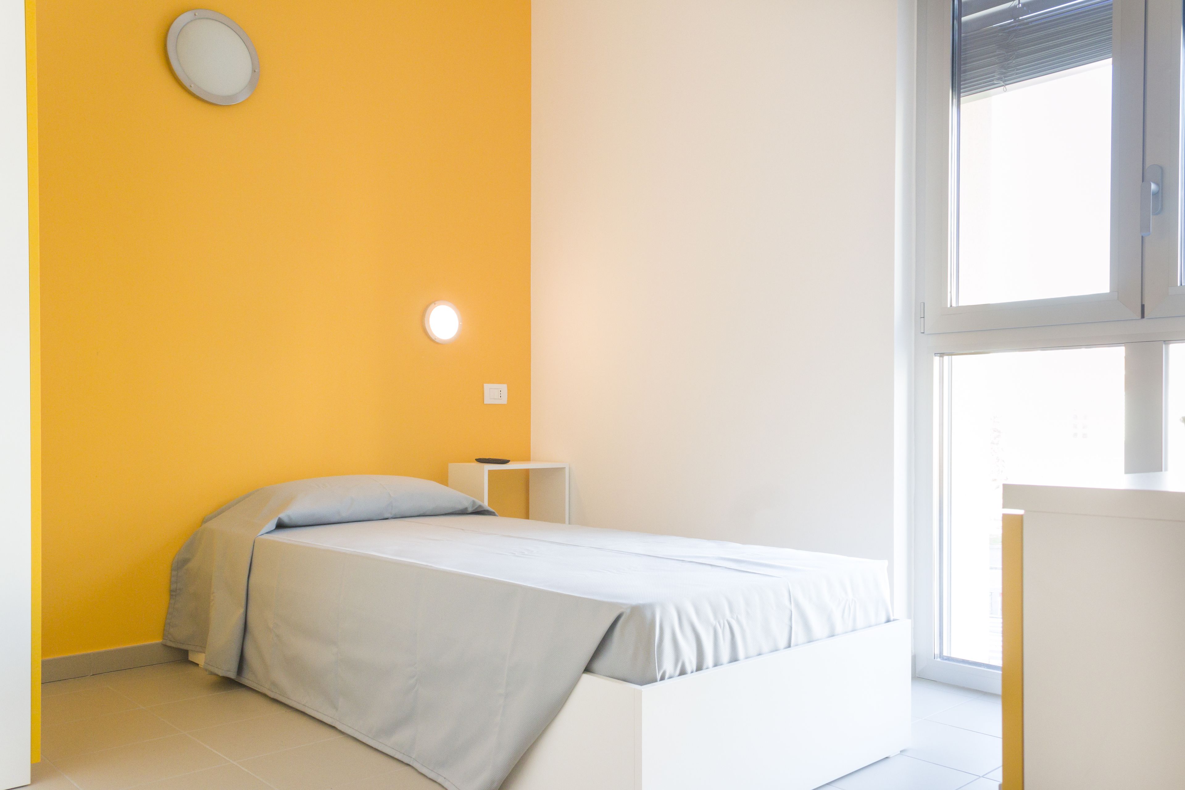 Quarto com cama, mesa de cabeceira, janela e luminária fixada na parede.