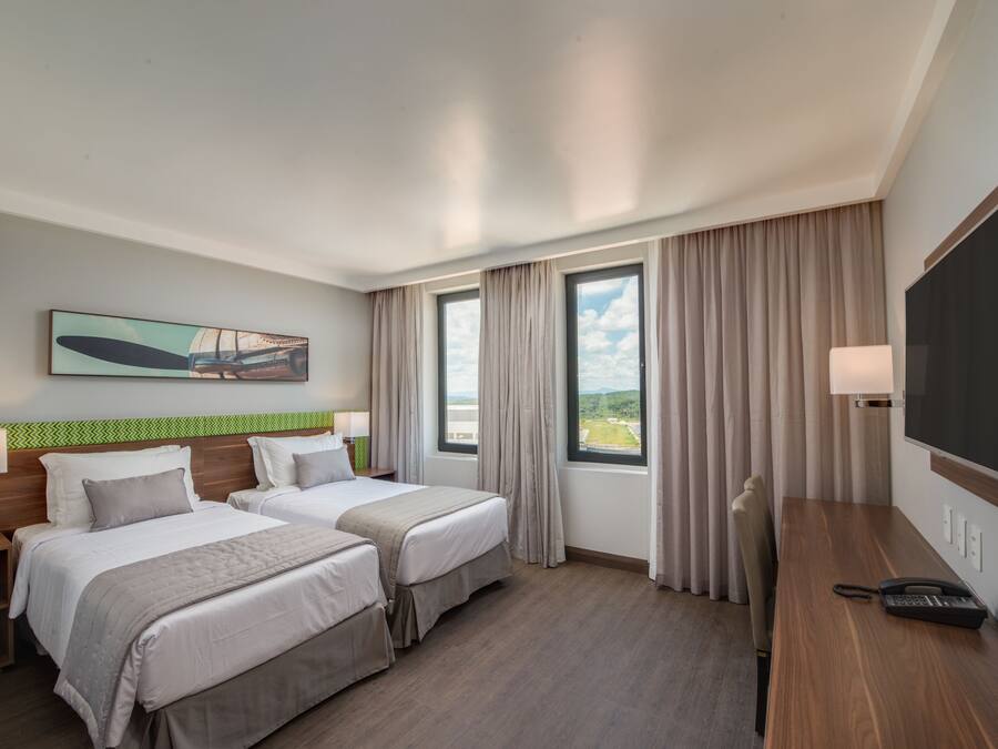 Quarto de hotel com duas camas, uma escrivaninha, televisão e janela com cortinas.