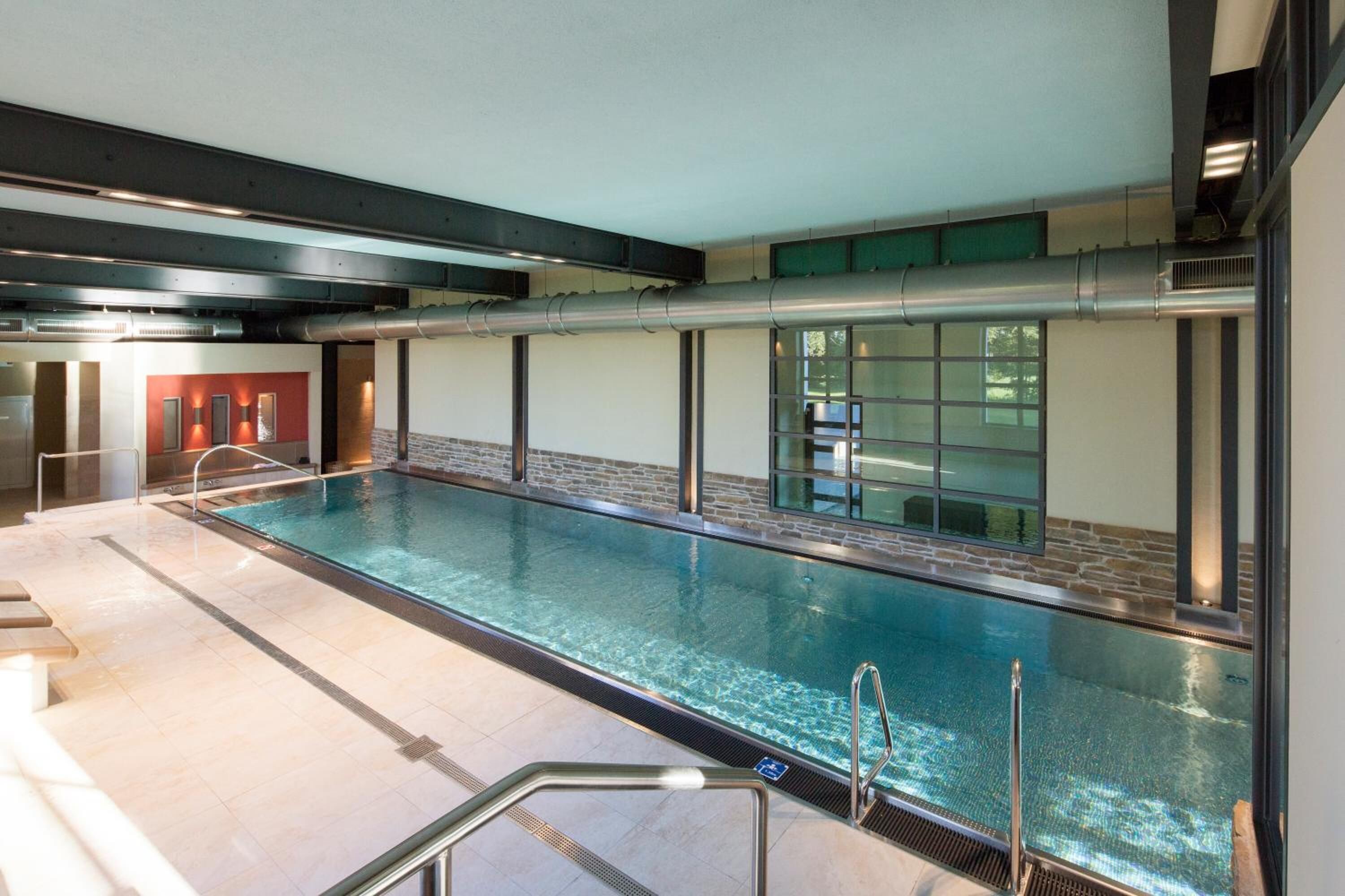 Piscina interna com água cristalina, piso cerâmico e uma parede de vidro de um lado.