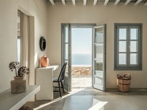 Quarto com parede de pedra, um banco branco, uma cadeira, uma cesta de vime e vista para o mar através de uma porta aberta.