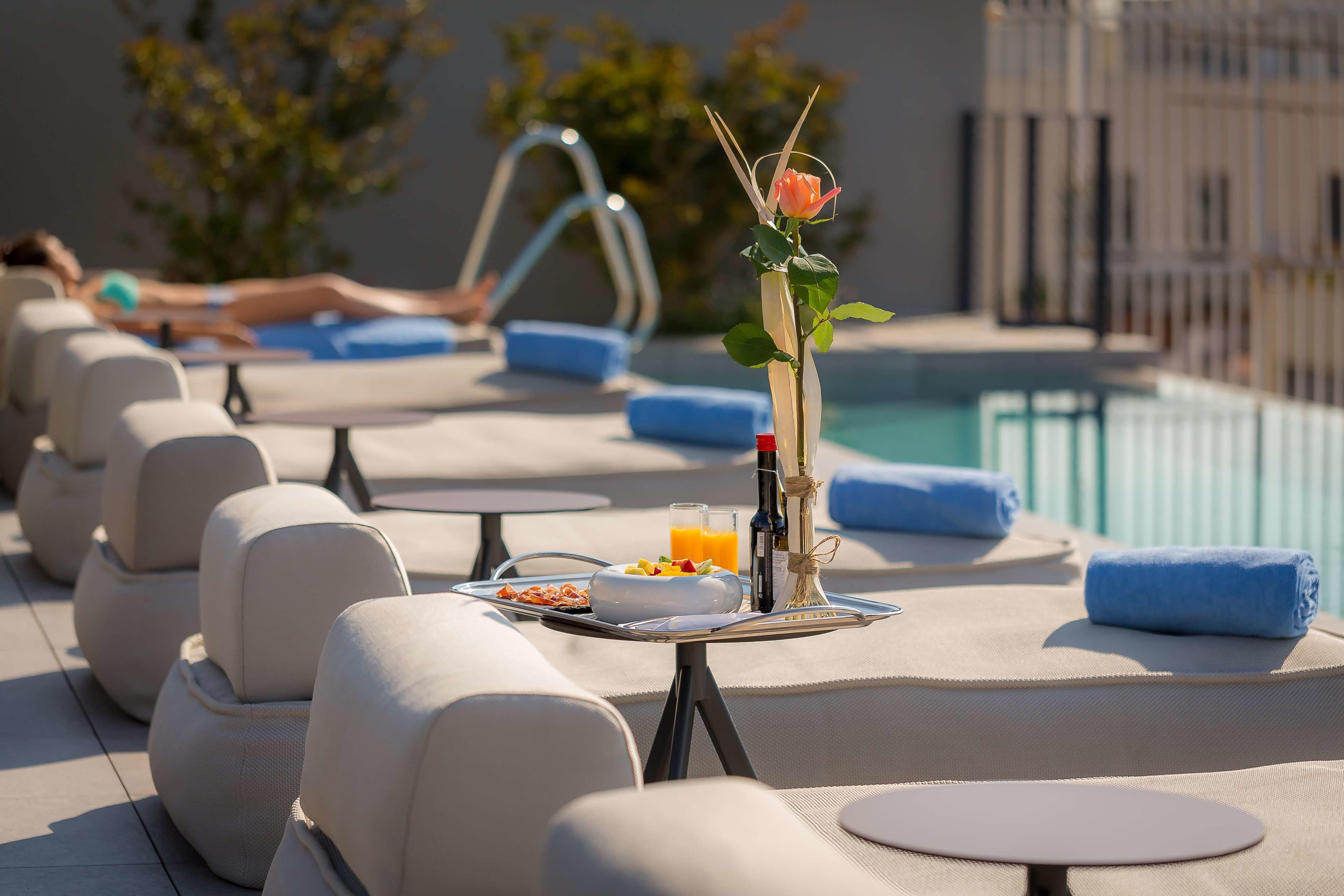 Uma área de estar à beira da piscina, com espreguiçadeiras, uma mesa com uma garrafa, copos e uma cesta de frutas.