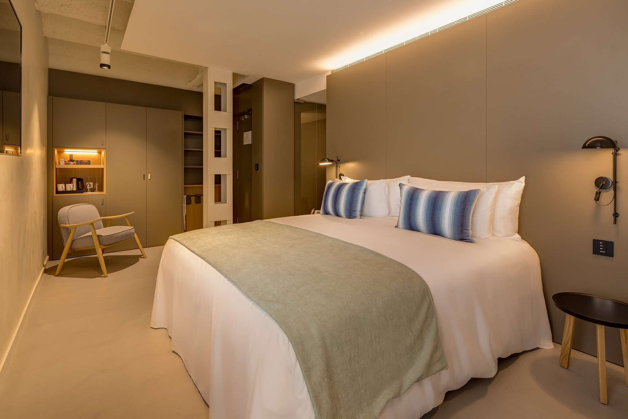 Quarto de hotel moderno com uma cama grande, uma poltrona, mesa de cabeceira e guarda-roupa embutido.