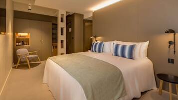 Quarto de hotel moderno com uma cama grande, uma poltrona, mesa de cabeceira e guarda-roupa embutido.