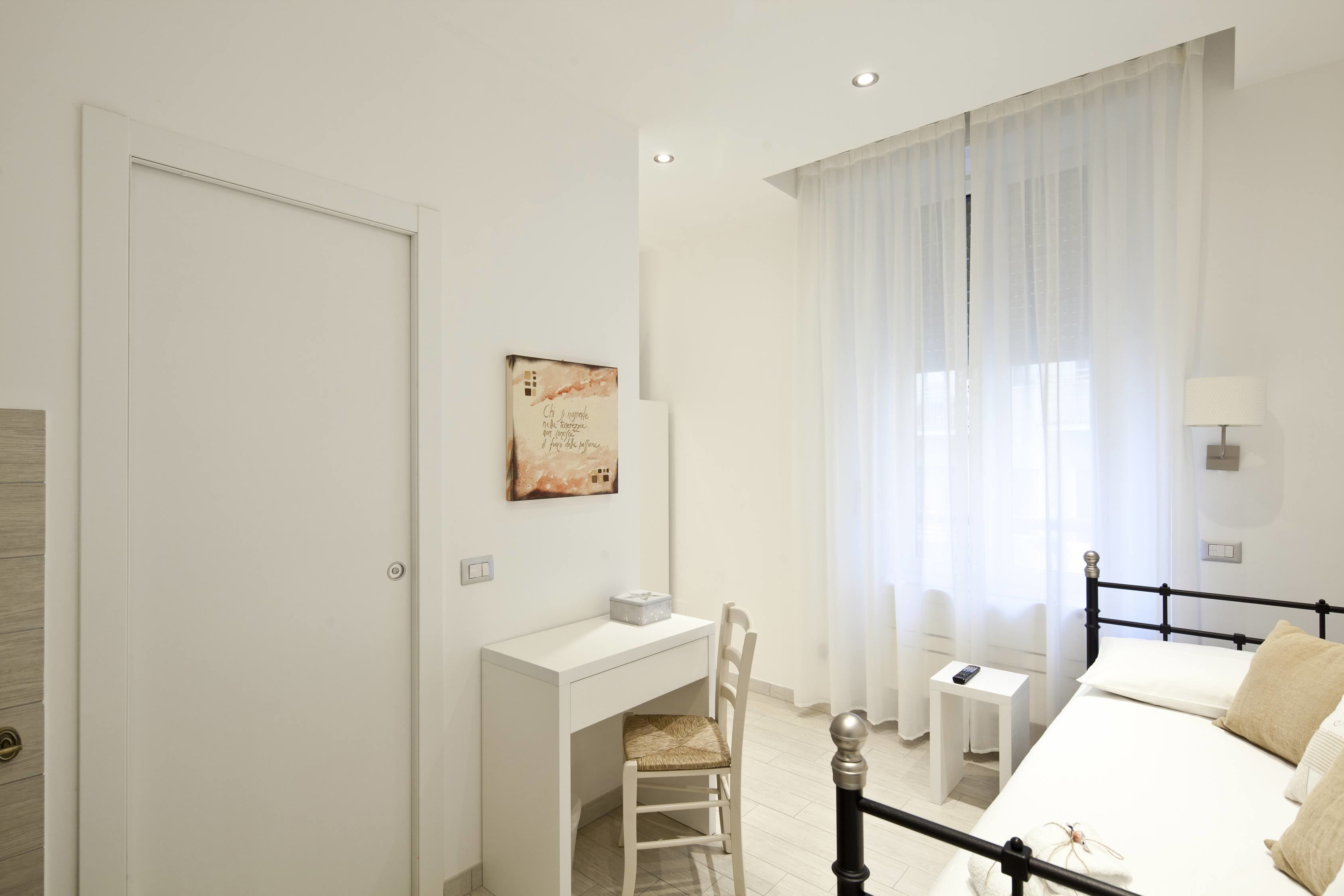 Quarto com cama, escrivaninha, cadeira, mesinha e janela com cortinas.