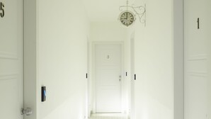 Um corredor claro e branco, com portas numeradas de 1 a 5, que dá para um quarto com um relógio na parede.
