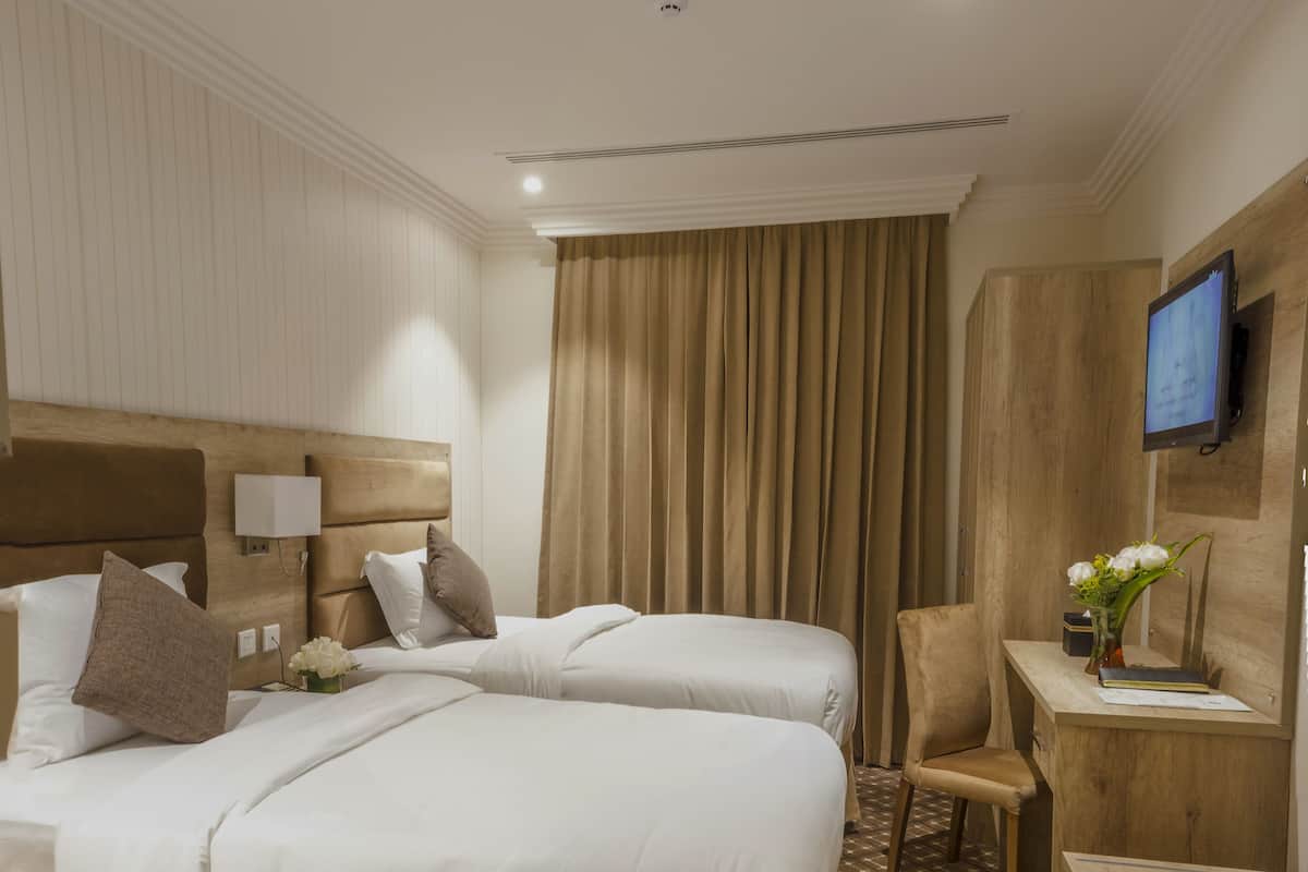 Quarto de hotel com duas camas, uma escrivaninha, uma cadeira, televisão e uma janela grande com cortinas.