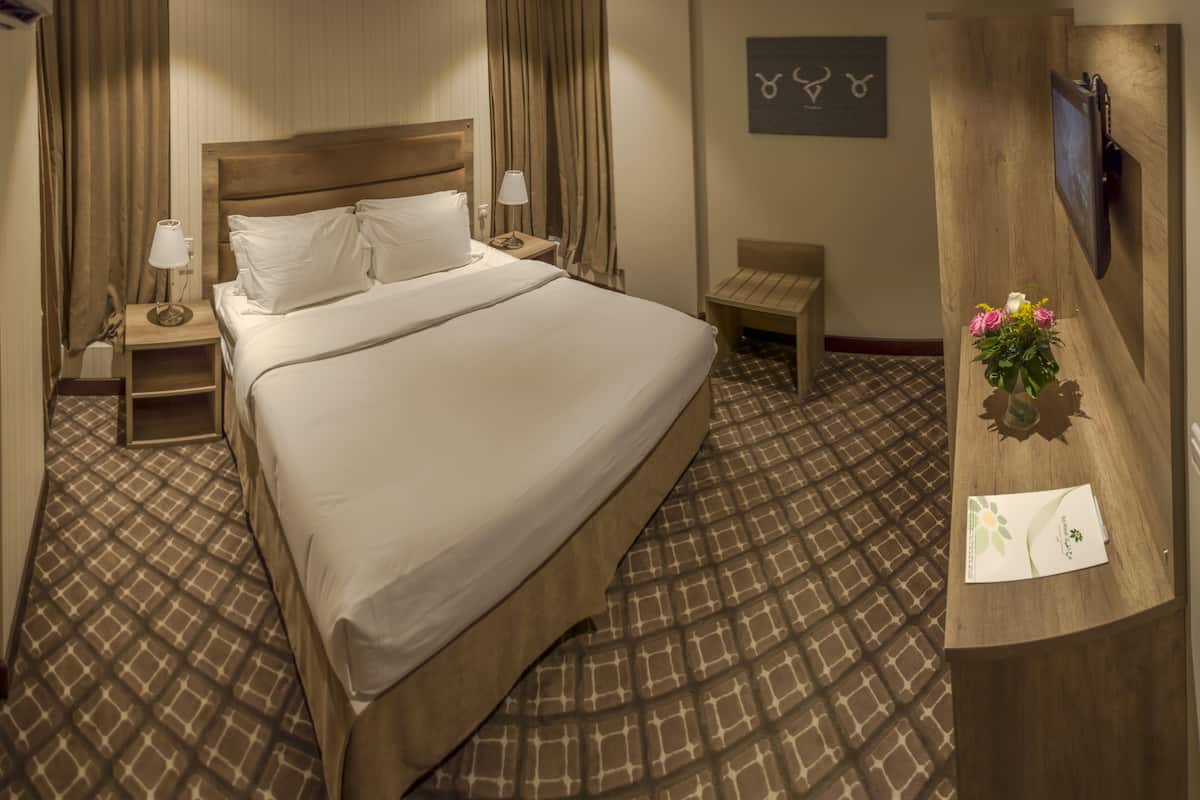 Quarto de hotel com uma cama grande, mesas de cabeceira, uma escrivaninha com um vaso de flores e uma TV fixada na parede.