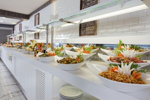 Buffet com diversos pratos, incluindo saladas e carnes, disposto sobre um balcão branco com vitrines de vidro.