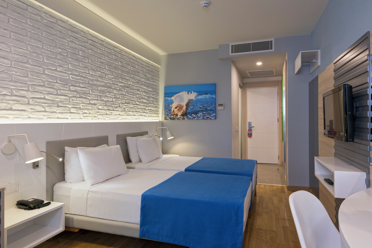 Quarto de hotel moderno com parede em padrão de tijolos, uma cama grande com roupa de cama branca e um toque de azul, uma escrivaninha com telefone, um espaço de estar com sofá branco e uma televisão montada.
