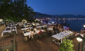 Uma área de refeições ao ar livre com mesas preparadas para hóspedes, com vista para um porto com barcos e um horizonte urbano ao fundo.