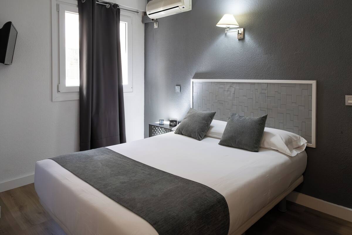 Uma cama bem arrumada com um cobertor cinza, uma cabeceira com padrão geométrico, uma mesa de cabeceira com abajur e uma janela com cortinas.