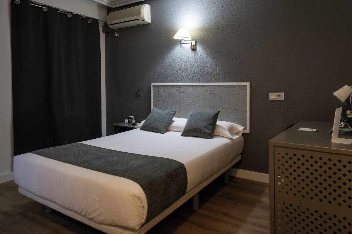 Quarto de hotel com cama, mesa de cabeceira, abajur e aparelho de ar condicionado.