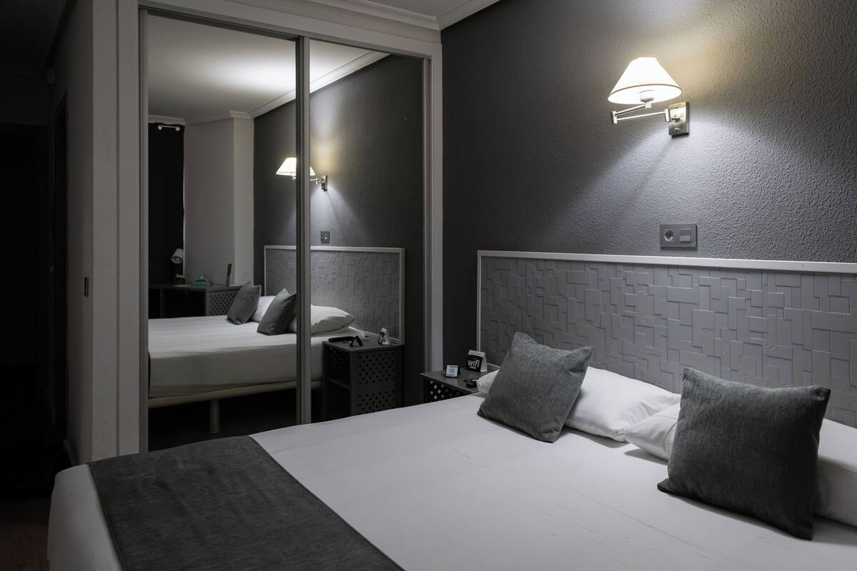Quarto de hotel com cama, mesa de cabeceira, luminária, espelho e parede com padrão geométrico.