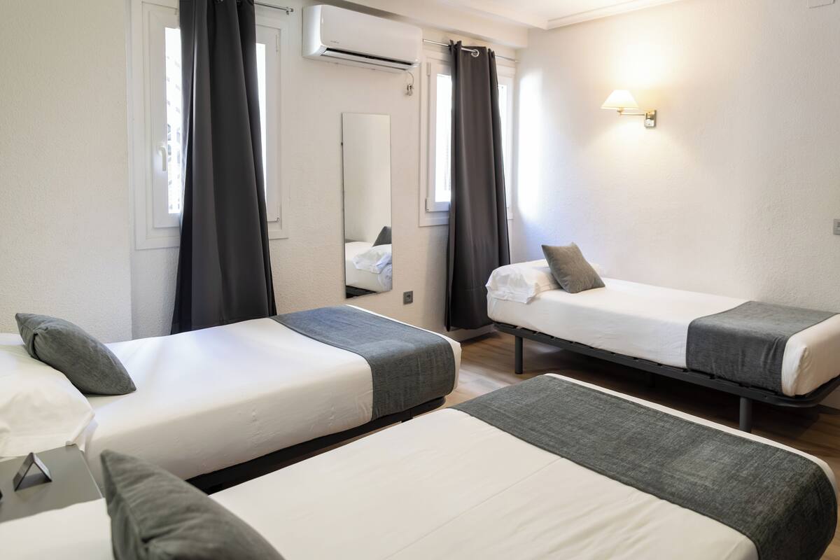 Quarto com duas camas de solteiro, um espelho, ar condicionado, janela com cortinas e uma luminária na parede.