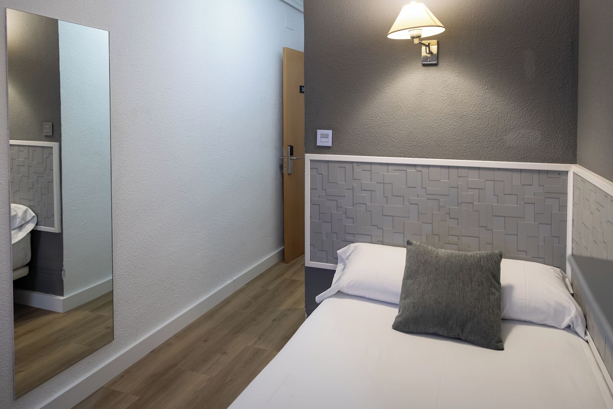Quarto de hotel com uma cama, abajur, espelho e porta.
