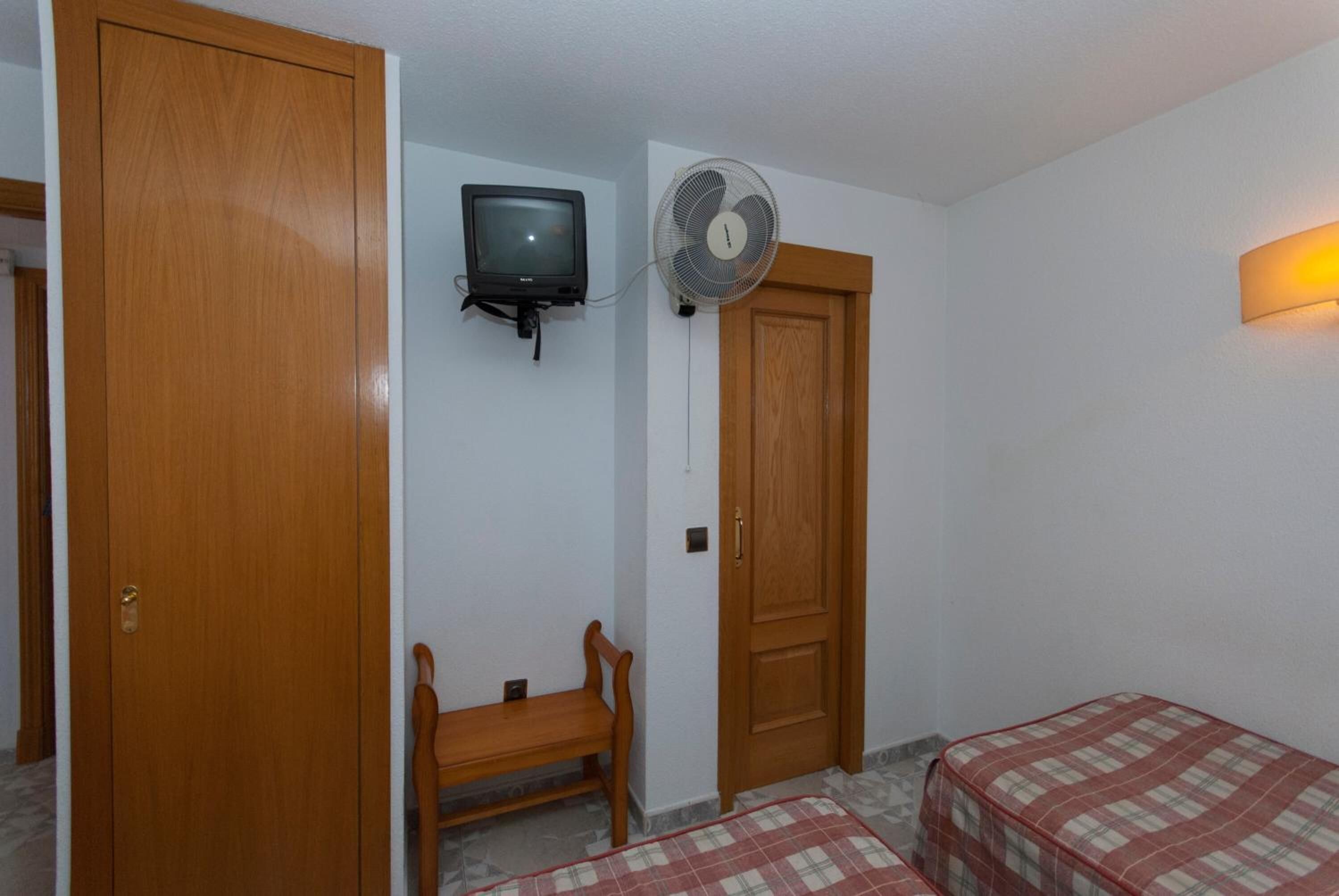 Um quarto com porta de madeira, televisão fixada na parede, ventilador portátil, um banco de madeira e uma cama com colcha xadrez.