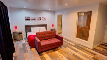 Quarto de hotel com cama, sofá vermelho, piso de madeira, mesa de cabeceira com abajur e parede decorada com quadros.