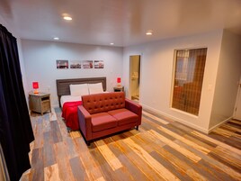Quarto de hotel com cama, sofá vermelho, piso de madeira, mesa de cabeceira com abajur e parede decorada com quadros.