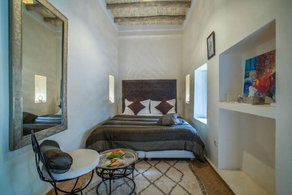 Quarto de hotel pequeno e estreito com uma cama, uma mesinha redonda com uma fruteira, uma cadeira, um espelho grande e um quadro na parede.
