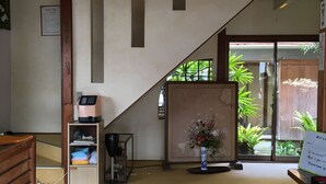 Um saguão de hotel com uma escada, um balcão de recepção, um telefone, uma máquina de vendas automática, uma pequena mesa com um vaso e uma placa.